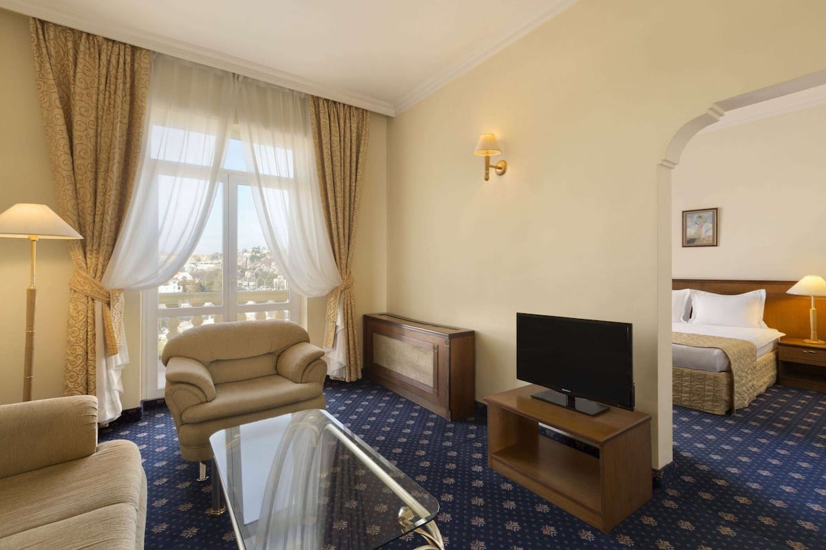 Quarto de hotel com sofá, televisão em móvel, cama com cabeceira e janela com cortinas.