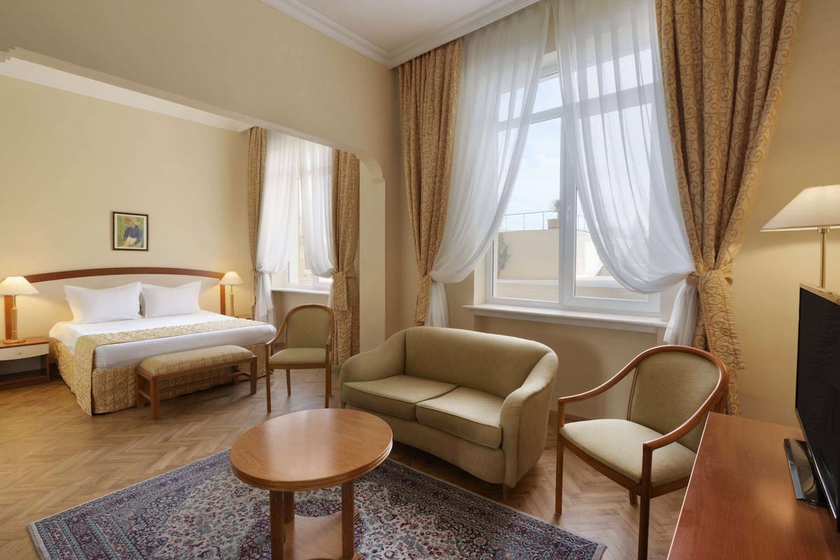 Quarto de hotel com uma cama, duas cadeiras, um sofá, uma mesinha e uma televisão.