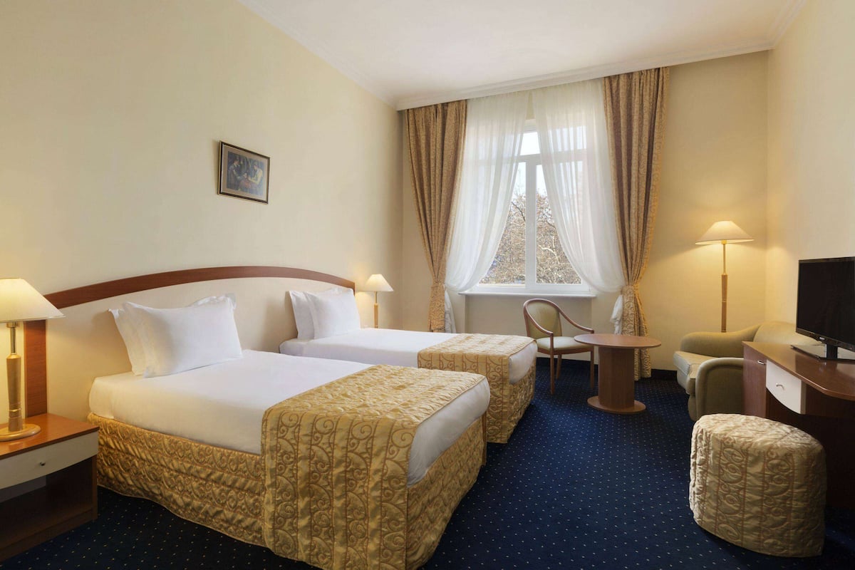 Quarto de hotel com duas camas, uma escrivaninha, uma cadeira, televisão e janela com cortinas.