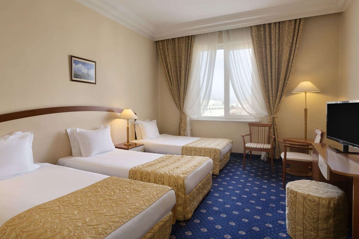 Quarto de hotel com três camas, uma escrivaninha com televisão, uma cadeira e uma janela com cortinas.