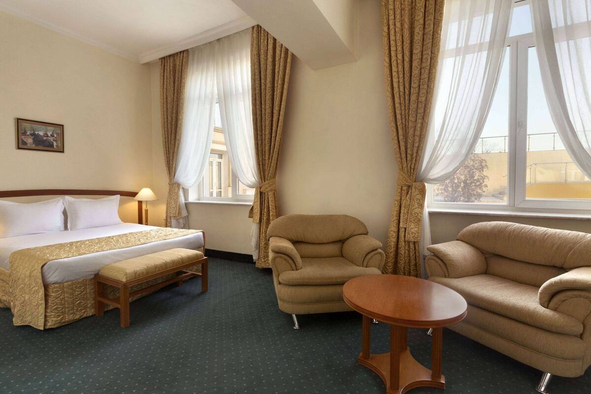 Quarto de hotel com cama, sofá, cadeira, mesa pequena e cortinas.