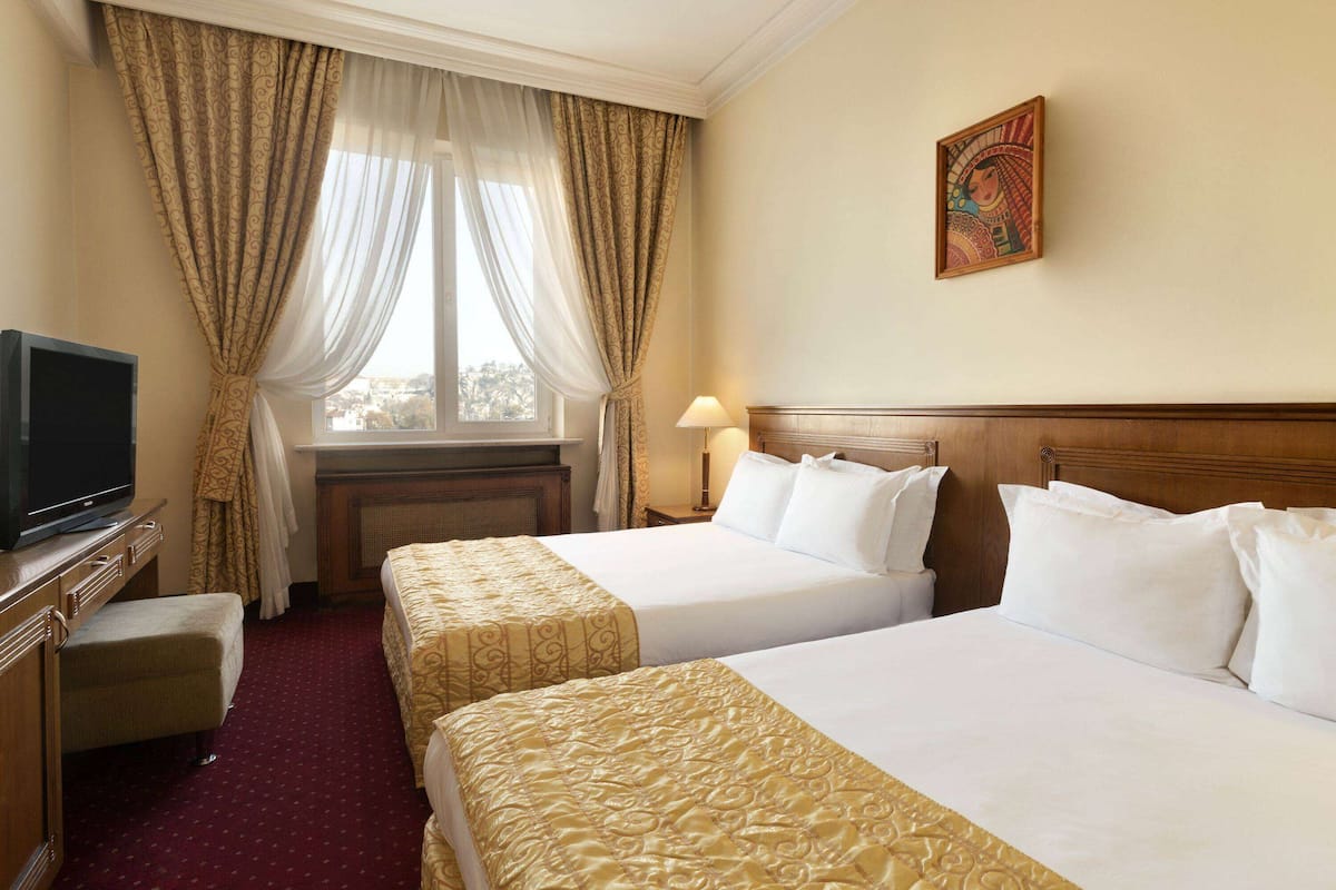 Quarto de hotel com duas camas, televisão, escrivaninha e janela com cortinas.