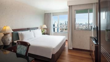 Quarto de hotel com uma cama grande, duas mesas de cabeceira com abajures, uma escrivaninha e vista para a cidade através de uma janela ampla.