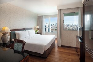 Quarto de hotel com uma cama grande, duas mesas de cabeceira com abajures, uma escrivaninha e vista para a cidade através de uma janela ampla.