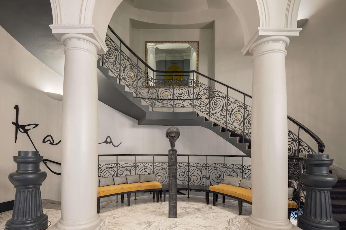Uma escadaria imponente com grades pretas ornamentadas, piso de mármore e um espaço com bancos e almofada amarela.