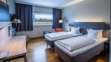 Quarto de hotel com duas camas, uma escrivaninha, uma cadeira, televisão e janela com cortinas.