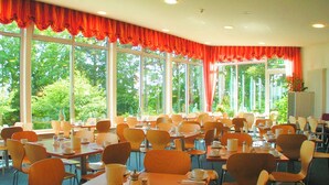 Uma sala de jantar com várias mesas e cadeiras, grandes janelas com cortinas vermelhas, e vista para o verde lá fora.