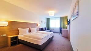 Quarto de hotel com uma cama grande, mesas de cabeceira, área de estar com sofá e poltronas, uma pequena escrivaninha e uma janela com cortinas.