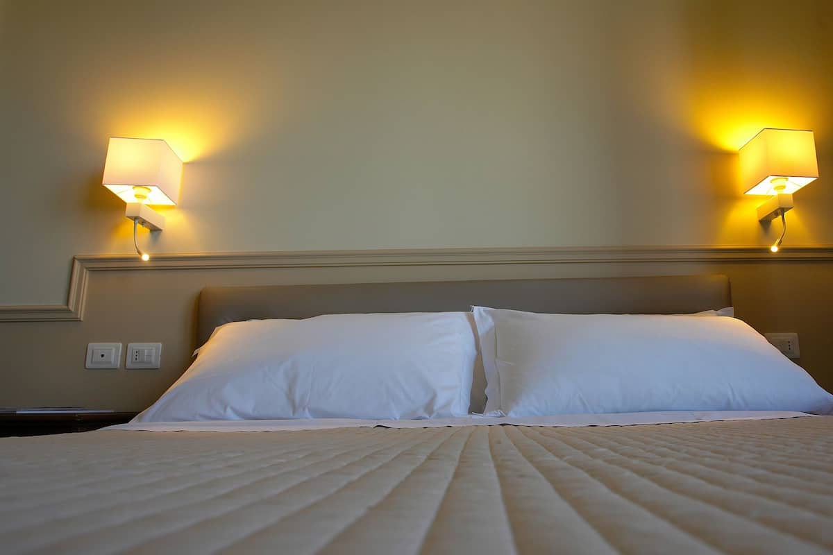 Cama bem arrumada com lençóis brancos e duas luminárias na parede, proporcionando iluminação suave.