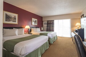 Quarto de hotel com duas camas, uma escrivaninha, uma cadeira e uma janela grande com cortinas.
