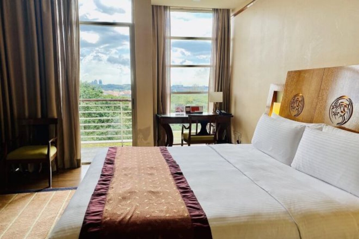 Quarto de hotel com uma cama grande, uma escrivaninha com abajur e vista para o verde lá fora.