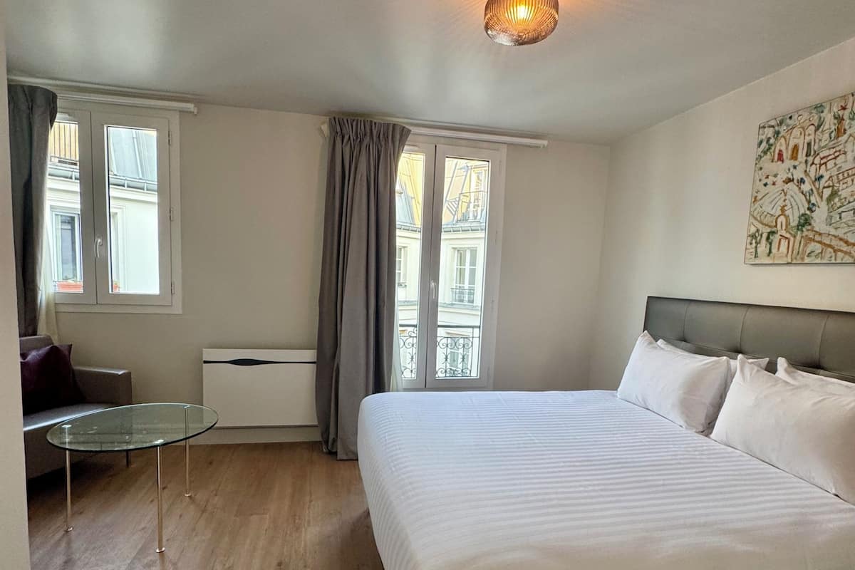 Quarto de hotel com cama, uma mesinha, sofá e janela com cortinas.