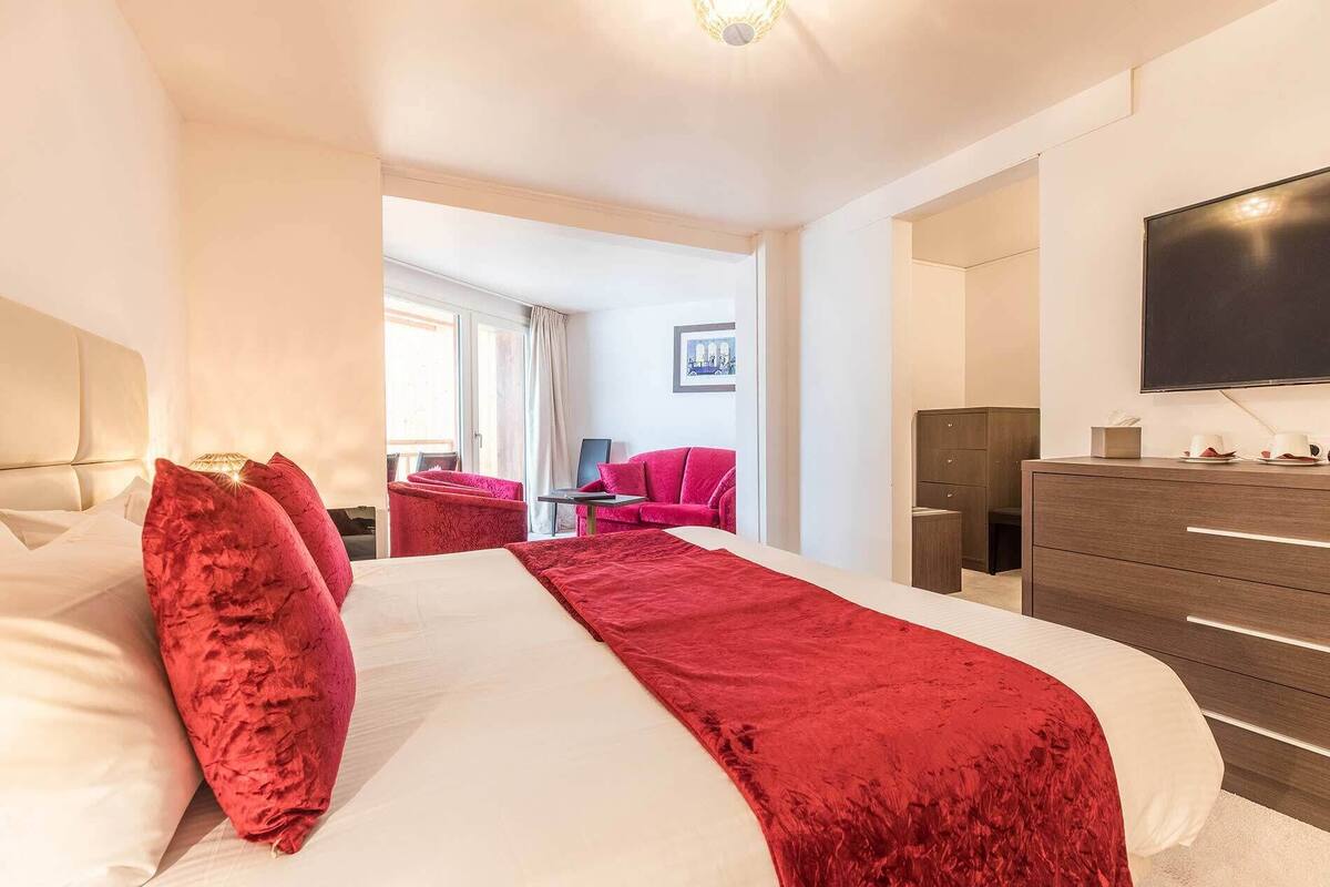 Quarto de hotel com uma cama grande, cômoda de madeira, televisão de tela plana e uma cadeira vermelha.