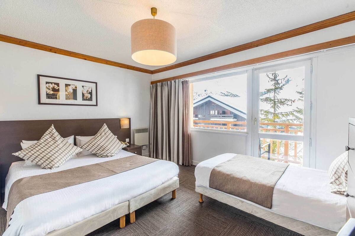 Quarto de hotel com duas camas, uma janela grande com vista para uma cabana de madeira e uma luminária no teto.