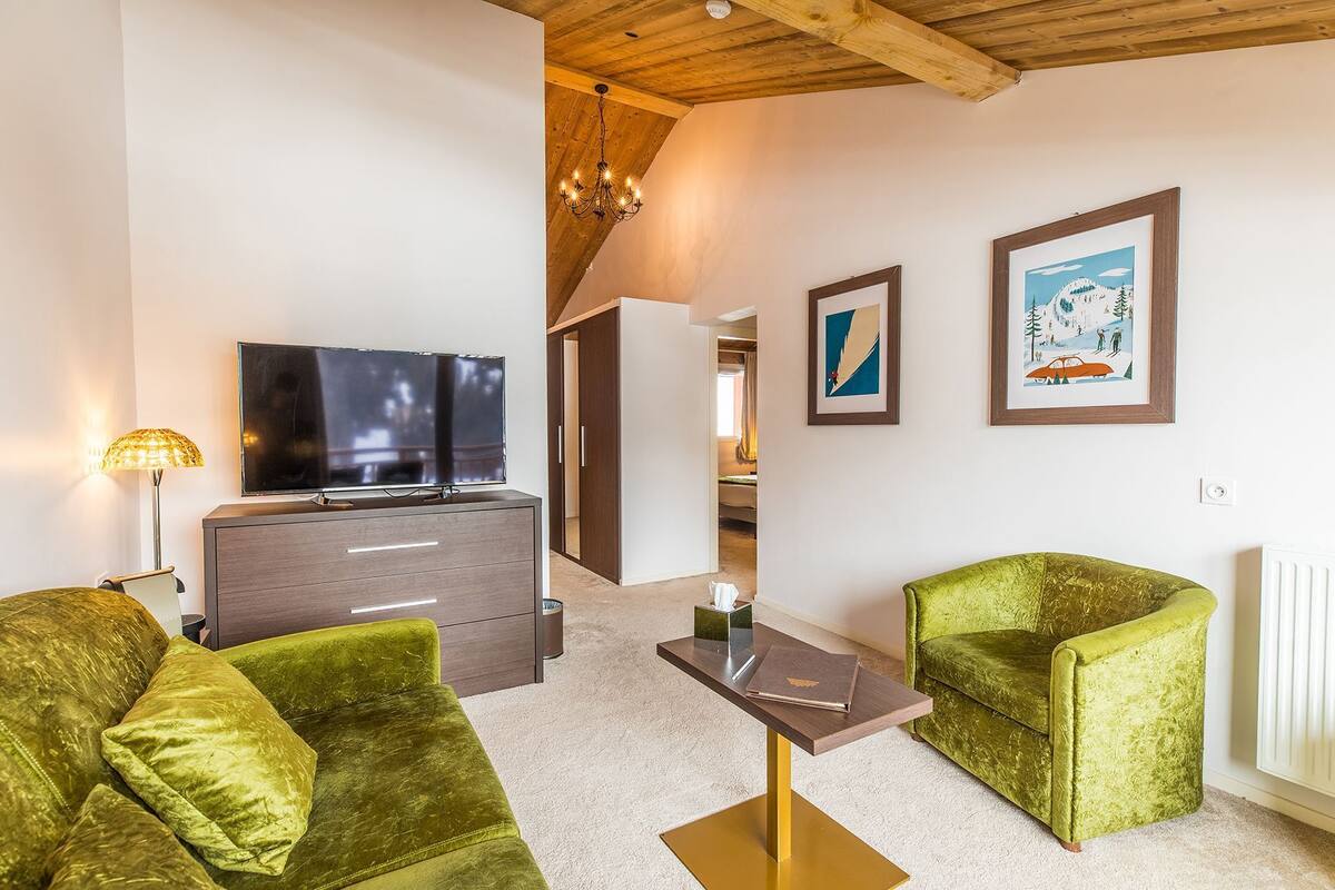 Uma sala de estar moderna com televisão, poltronas de veludo verde, um aparador de madeira e duas molduras na parede.