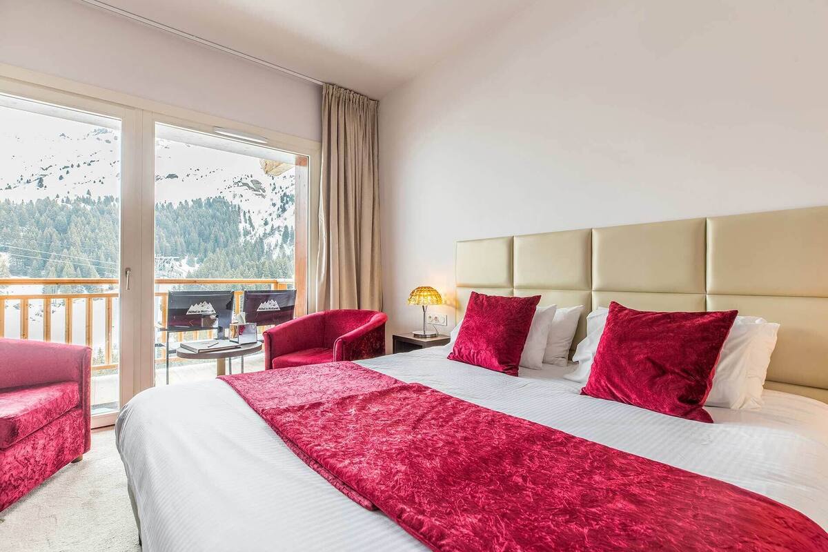 Quarto de hotel com cama, almofadas vermelhas, mesa de cabeceira, luminária, cadeira e varanda com vista para as montanhas.