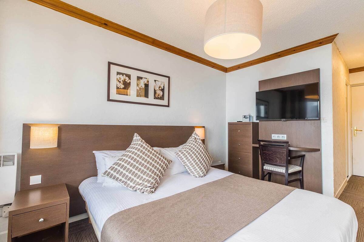 Quarto de hotel com cama, luminárias de cabeceira, escrivaninha com cadeira, televisão e um quadro na parede.