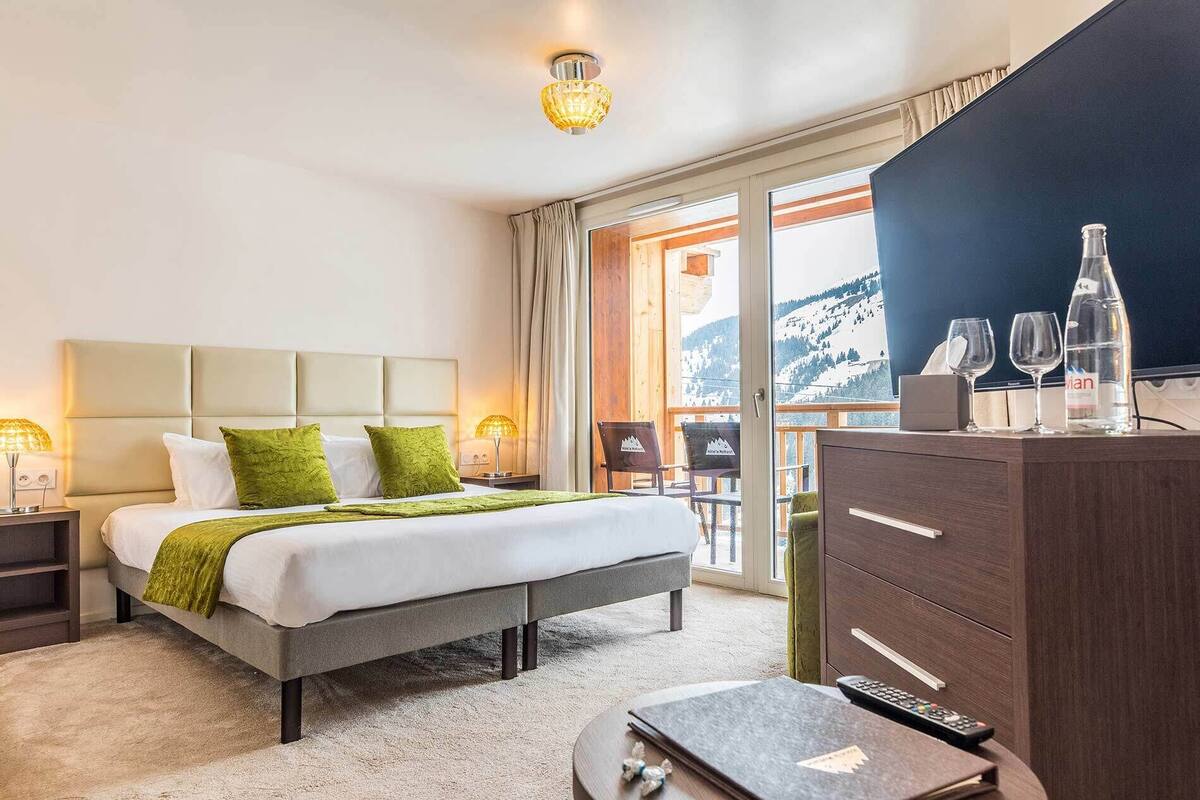 Quarto de hotel com cama, escrivaninha, penteadeira e varanda com vista para as montanhas.