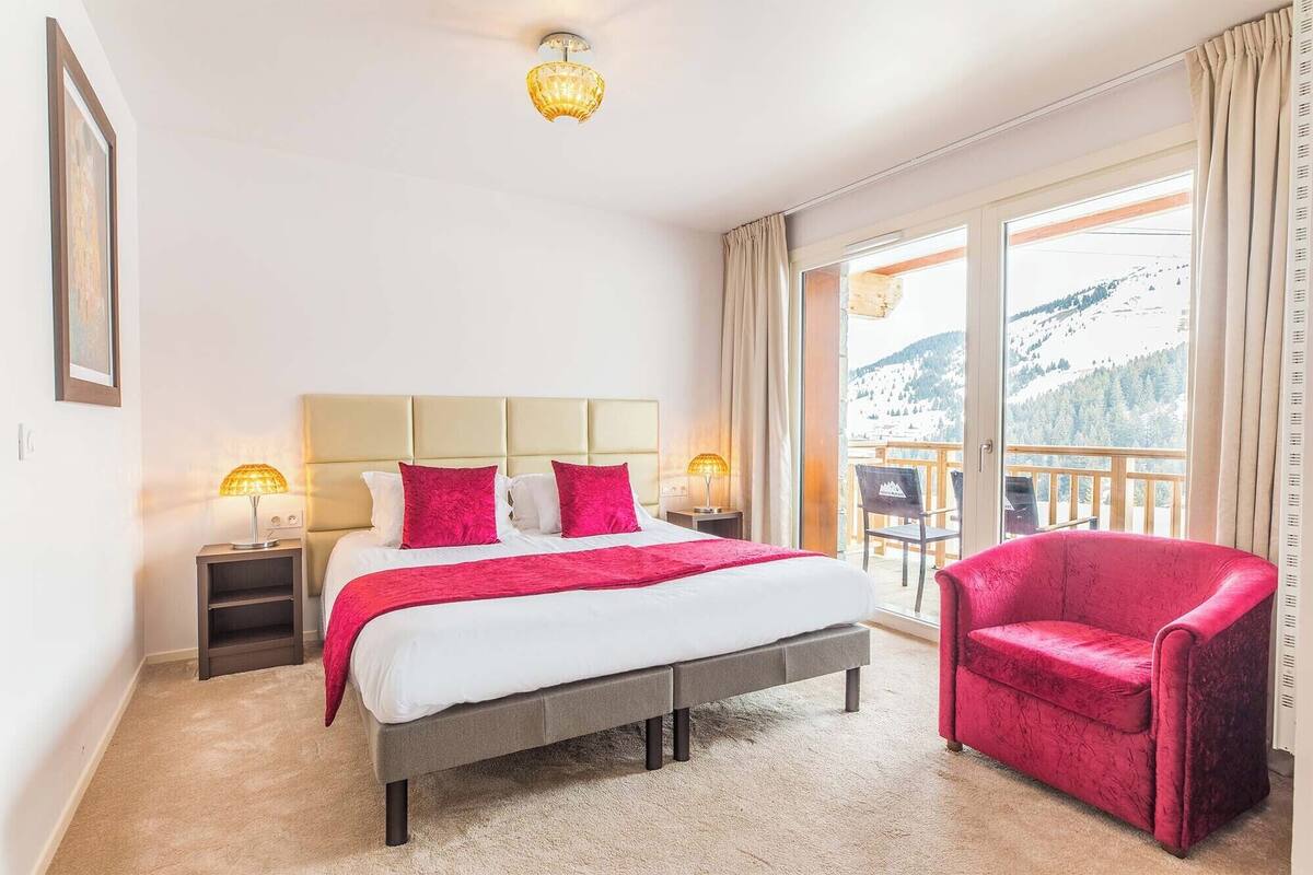 Quarto de hotel com uma cama, duas luminárias de cabeceira, uma cadeira vermelha e uma varanda com vista para uma montanha.