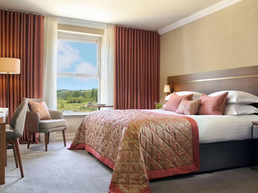 Quarto de hotel com uma cama grande, uma escrivaninha com luminária, uma cadeira e vista para uma paisagem verde pela janela.