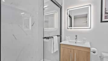 Um banheiro moderno com box de vidro, toucador de madeira, espelho com iluminação embutida e vaso sanitário.