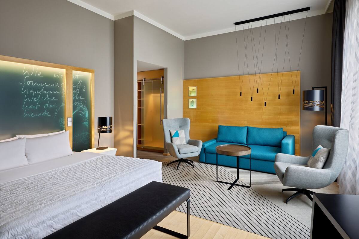 Quarto de hotel moderno com uma cama grande, duas poltronas, um sofá azul, uma mesa pequena e uma parede de destaque em madeira com um quadro negro.