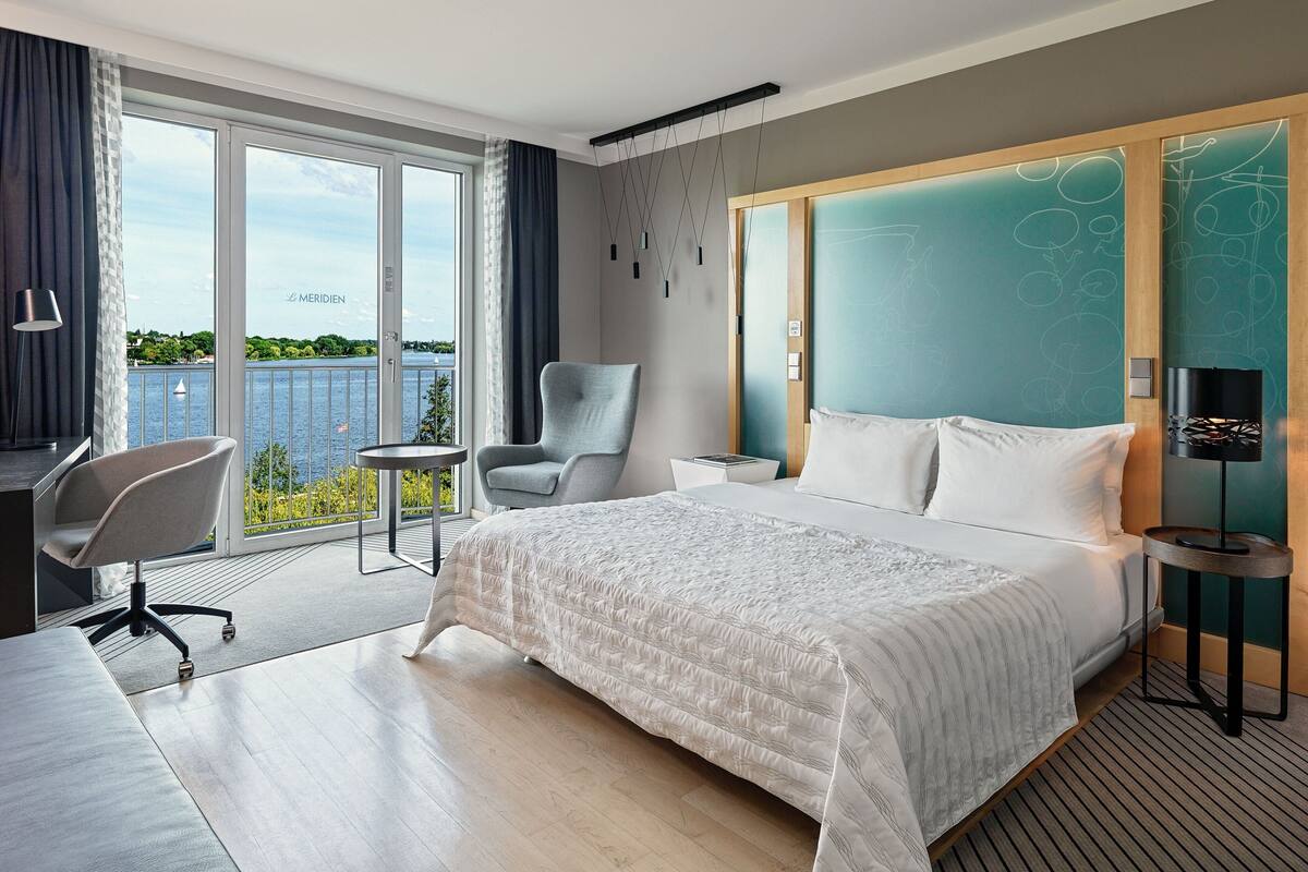 Quarto de hotel com uma cama grande, uma escrivaninha, uma cadeira, uma mesinha e vista para a água.