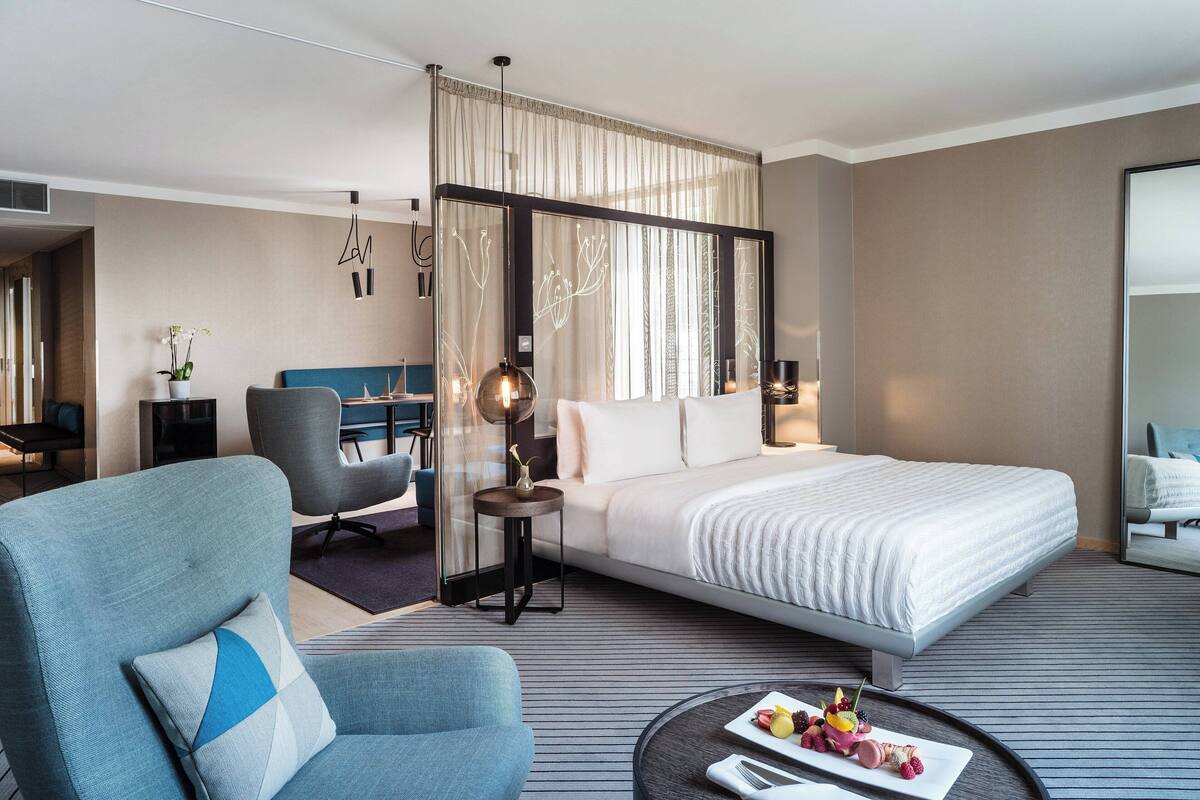 Quarto de hotel moderno com uma cama grande, área de estar com uma poltrona azul, uma pequena mesa redonda com um prato de comida e um espelho grande.