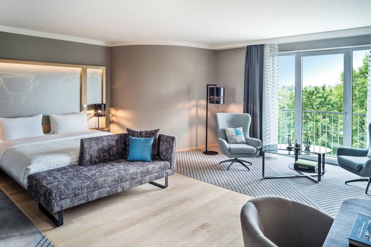 Quarto de hotel moderno com uma cama bem arrumada, um sofá, uma cadeira e uma mesa pequena. O quarto possui janelas amplas com vista para o verde lá fora.