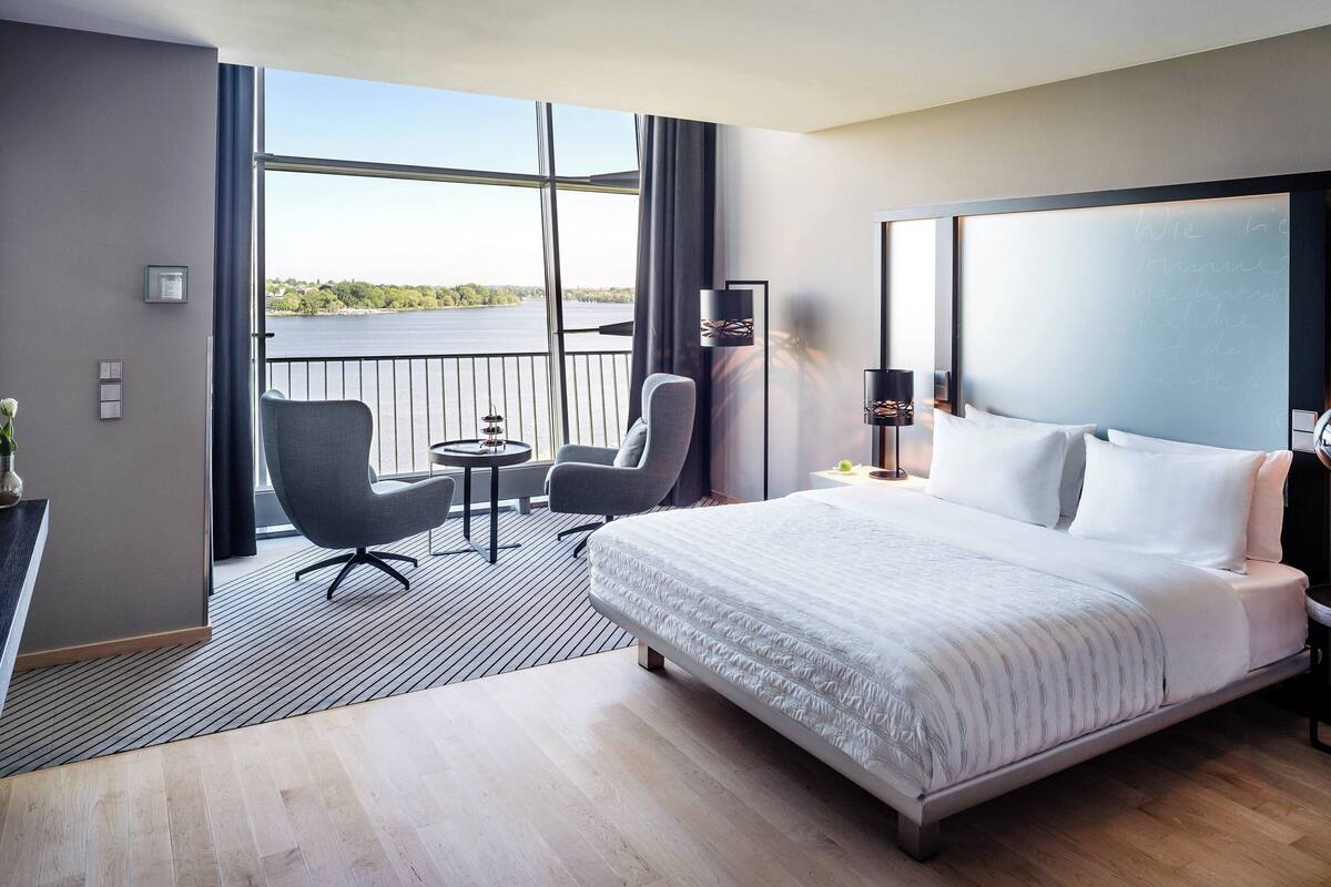 Quarto de hotel moderno com uma cama grande, uma mesa, uma cadeira e vista para um corpo de água através de grandes janelas.