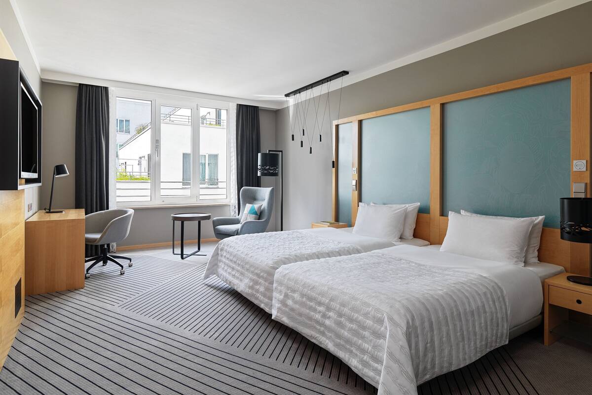 Quarto de hotel com duas camas, uma escrivaninha, uma cadeira e uma janela grande.