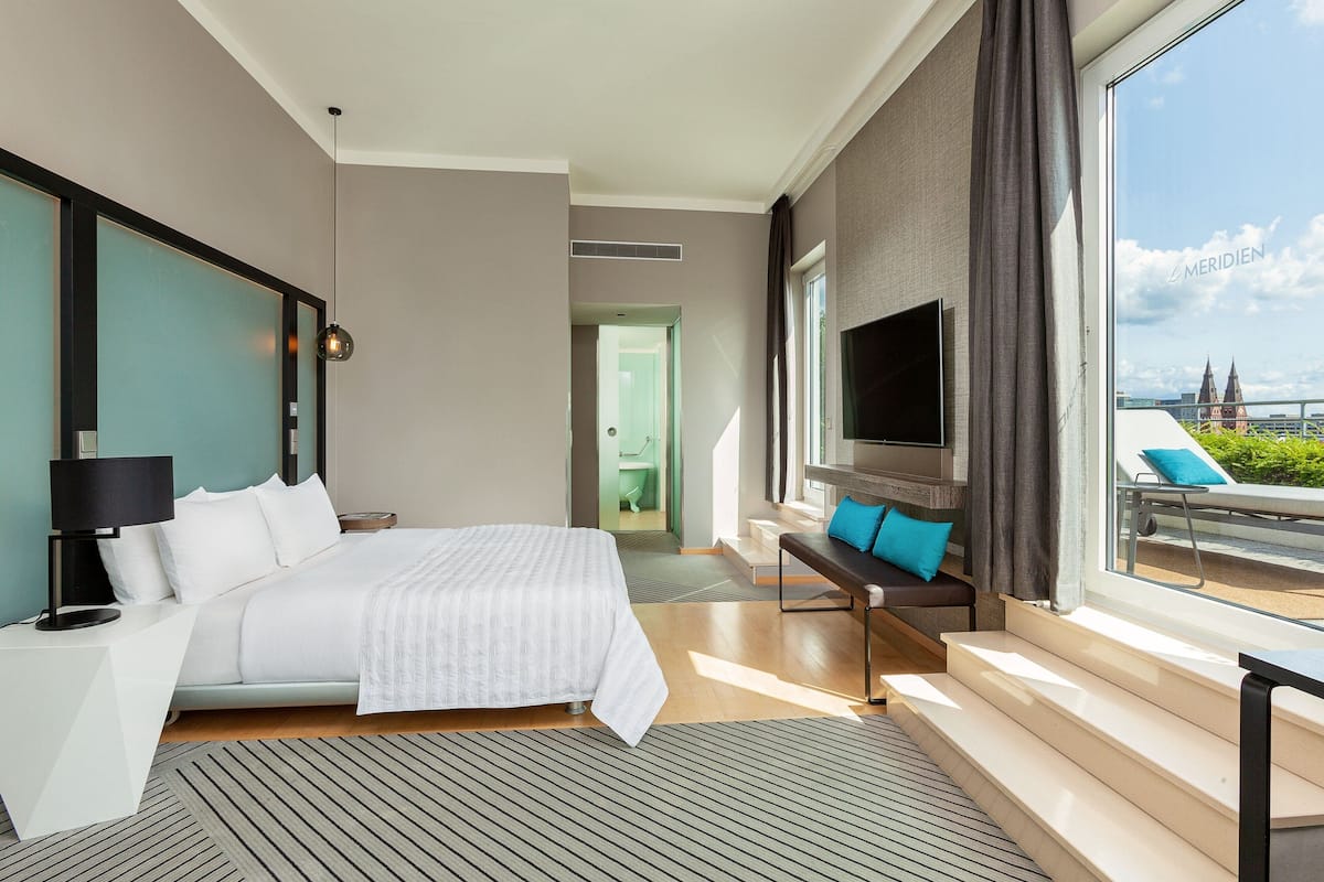 Quarto de hotel moderno com uma cama grande, televisão, varanda com vista e área de estar com almofadas.
