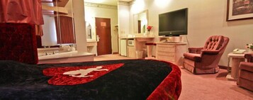 Quarto de hotel com cama, televisão, poltrona, escrivaninha e banheiro.
