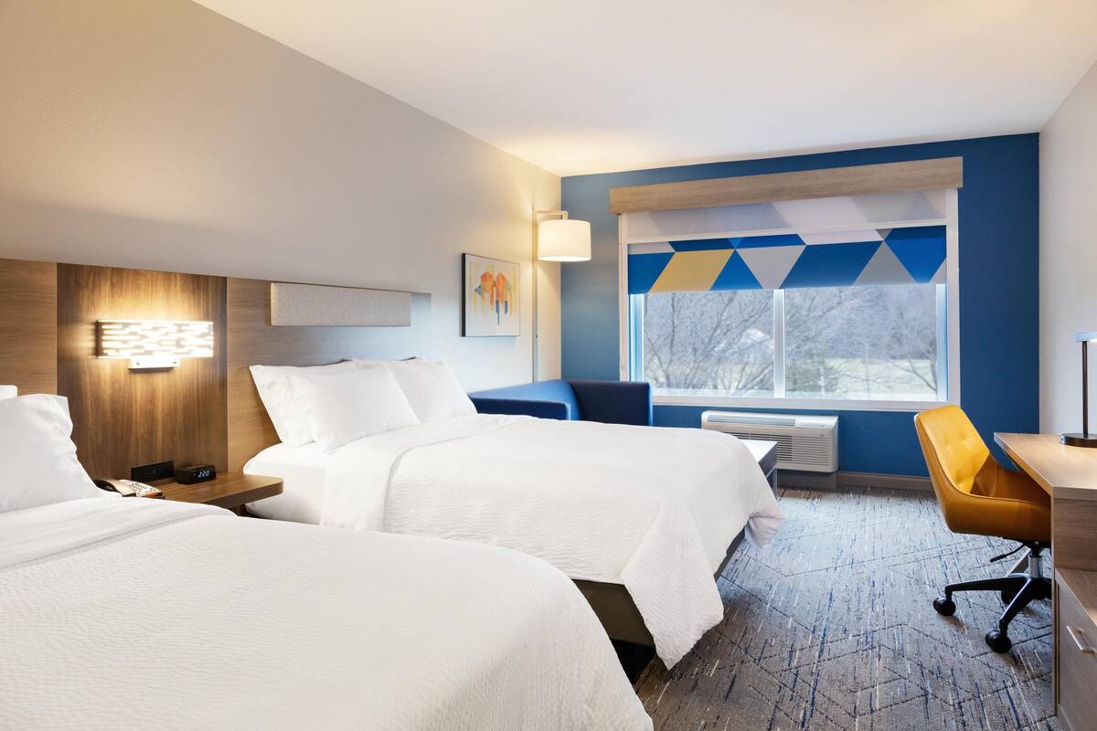 Quarto de hotel com duas camas, uma escrivaninha, uma cadeira, janela com vista e luminária na parede.