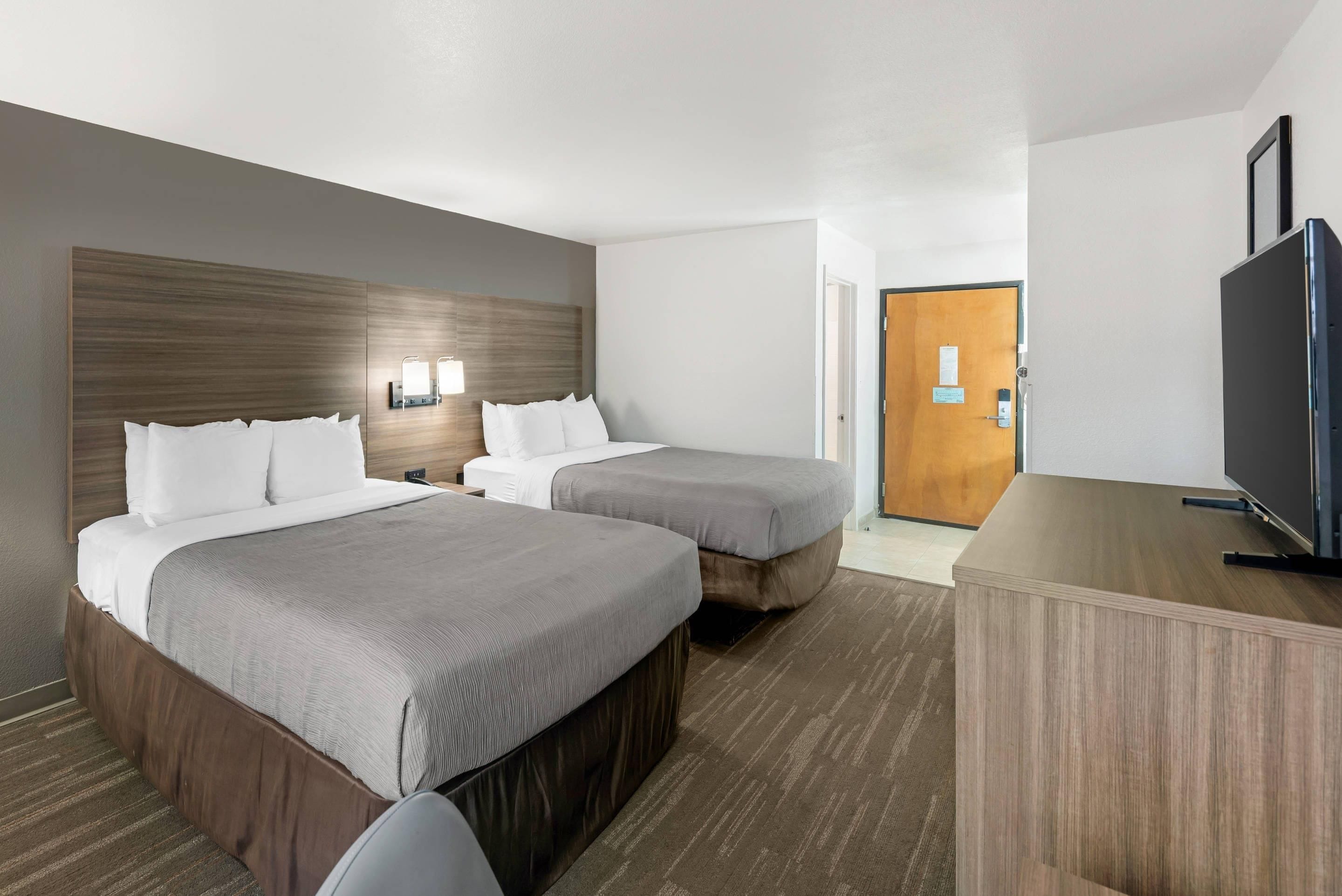Quarto de hotel com duas camas, uma escrivaninha, uma televisão e uma porta que dá para outro espaço.