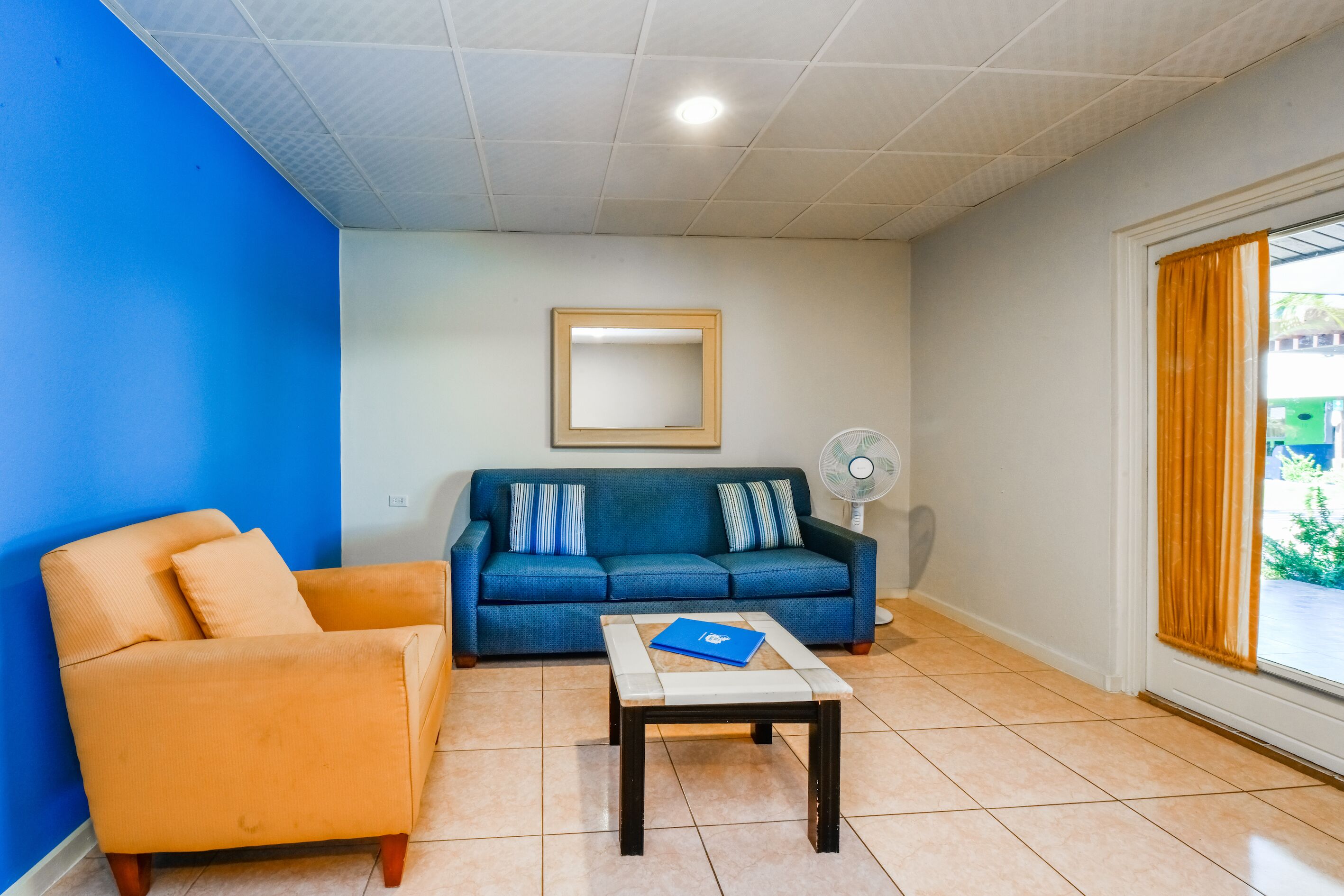 Uma sala de estar com um sofá azul, uma poltrona laranja, uma mesinha com uma pasta azul, um ventilador e uma janela com cortinas.