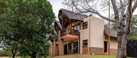 Uma casa com varanda, uma grande árvore no quintal e fachada de tijolos.