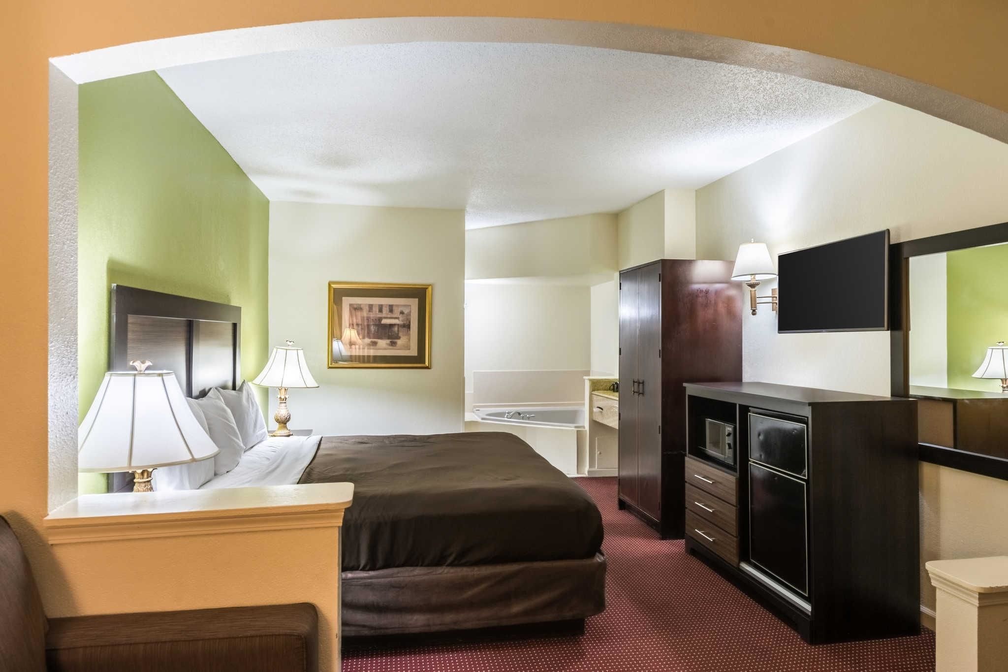 Quarto de hotel com cama, escrivaninha com luminária, cômoda, televisão e um quadro na parede.