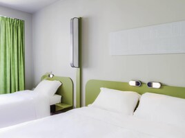 Quarto de hotel com duas camas, cabeceira verde, mesa de cabeceira com abajur e luz acoplada à parede.