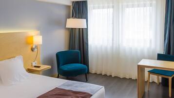 Quarto de hotel com cama, escrivaninha, cadeira, abajur e janela com cortinas.