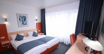 Quarto de hotel com cama, escrivaninha, cadeira, televisão e janela com cortinas.
