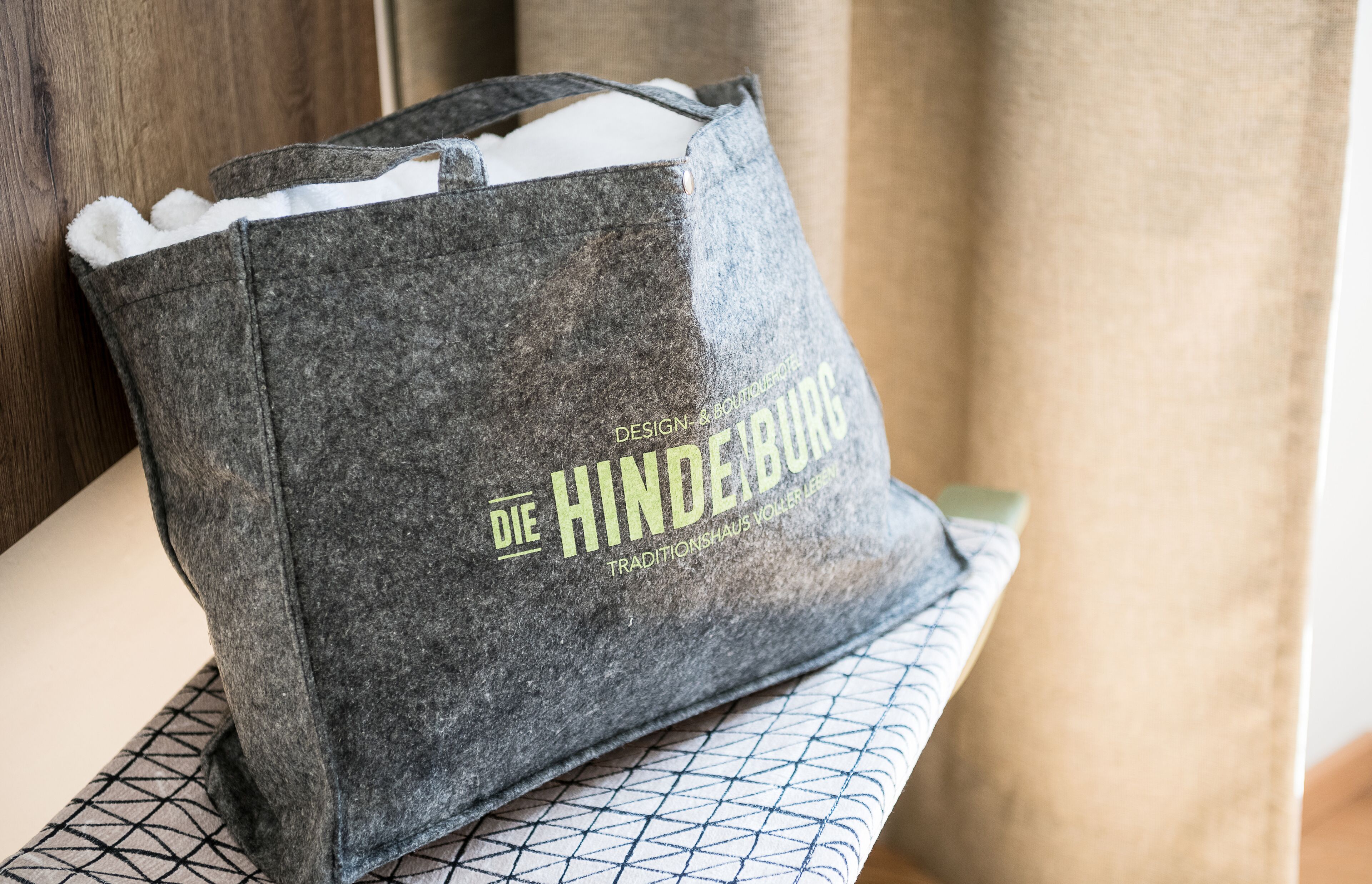 Uma bolsa de feltro cinza com o texto "DESIGN & CONCEPTS DIE HINDERUNG TRADITIONSHAUS VON KLEINER LEBEN" sobre uma superfície estampada.