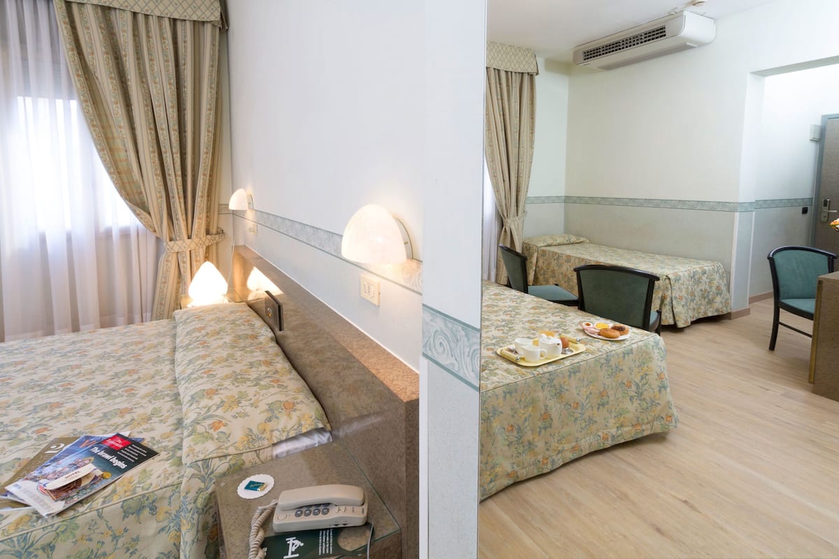 Quarto de hotel com cama, mesa de cabeceira com abajur, uma cadeira e pequena mesa com itens de café da manhã.