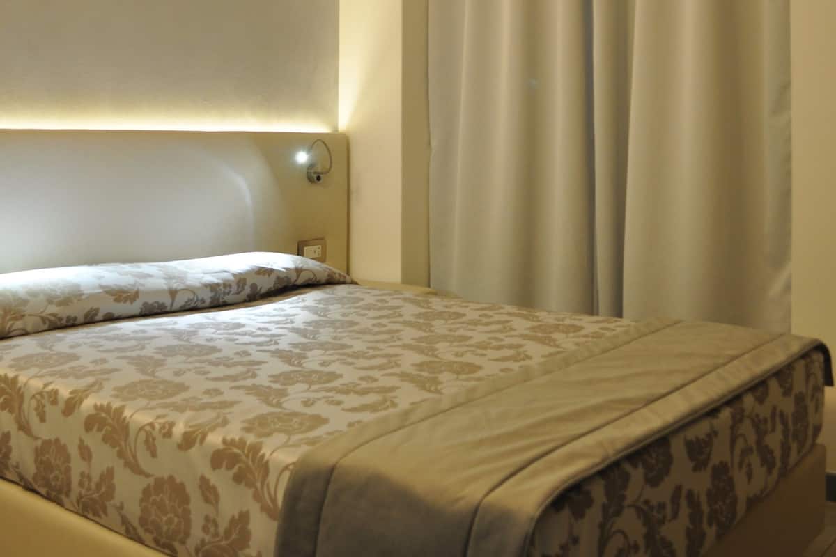 Quarto com cama, cabeceira, mesa de cabeceira, abajur e janela com cortinas.