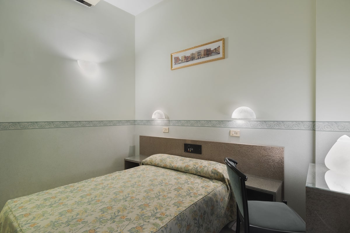 Quarto de hotel com cama, uma cadeira, escrivaninha, um quadro na parede e duas luminárias de parede.