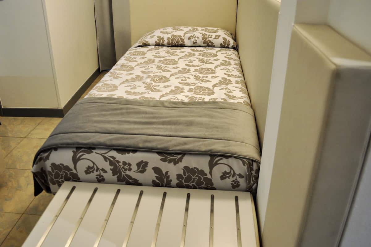 Quarto de hotel pequeno e estreito, com uma cama de solteiro, uma mesinha e um radiador.
