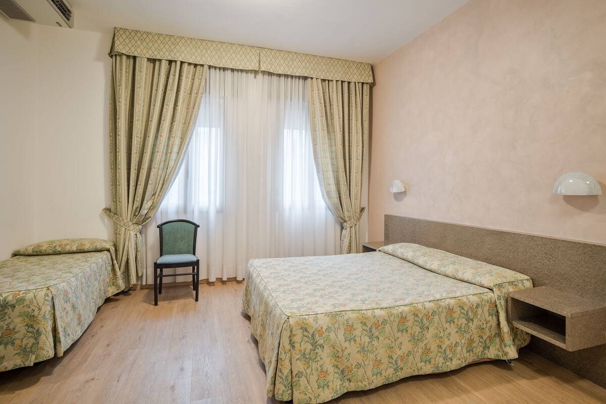 Quarto de hotel com duas camas de solteiro, uma cadeira, uma mesa de cabeceira e uma janela com cortinas.