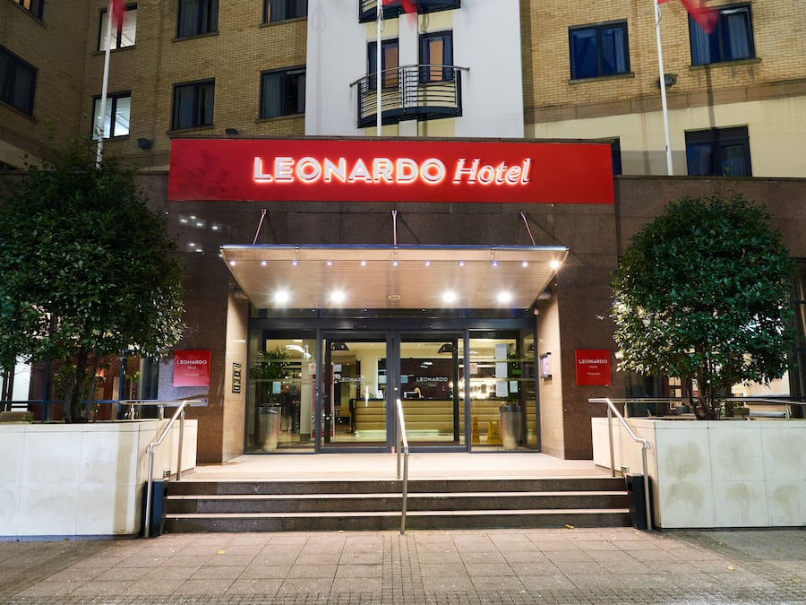 Exterior do Hotel Leonardo, com uma placa vermelha em destaque, um conjunto de degraus levando à entrada e duas árvores emoldurando a entrada.