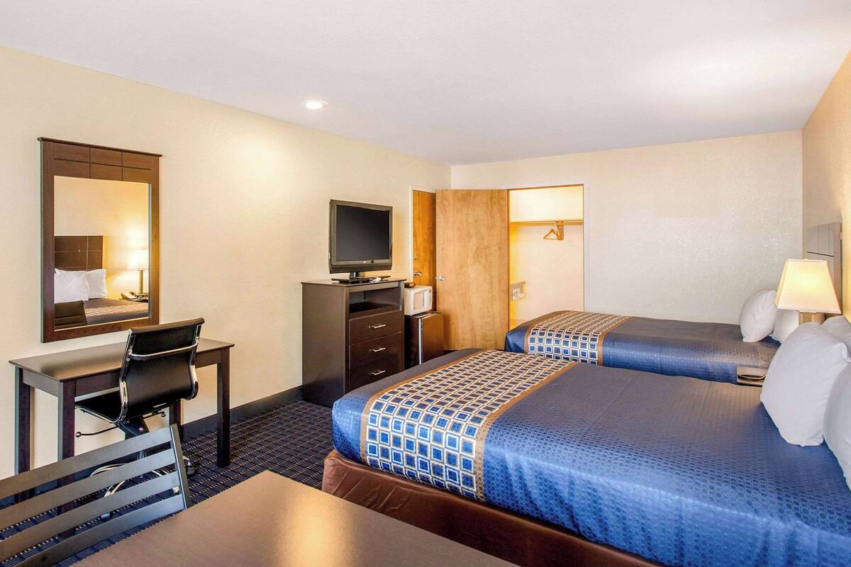Quarto de hotel com duas camas, uma escrivaninha, uma cadeira, uma televisão e um espelho.