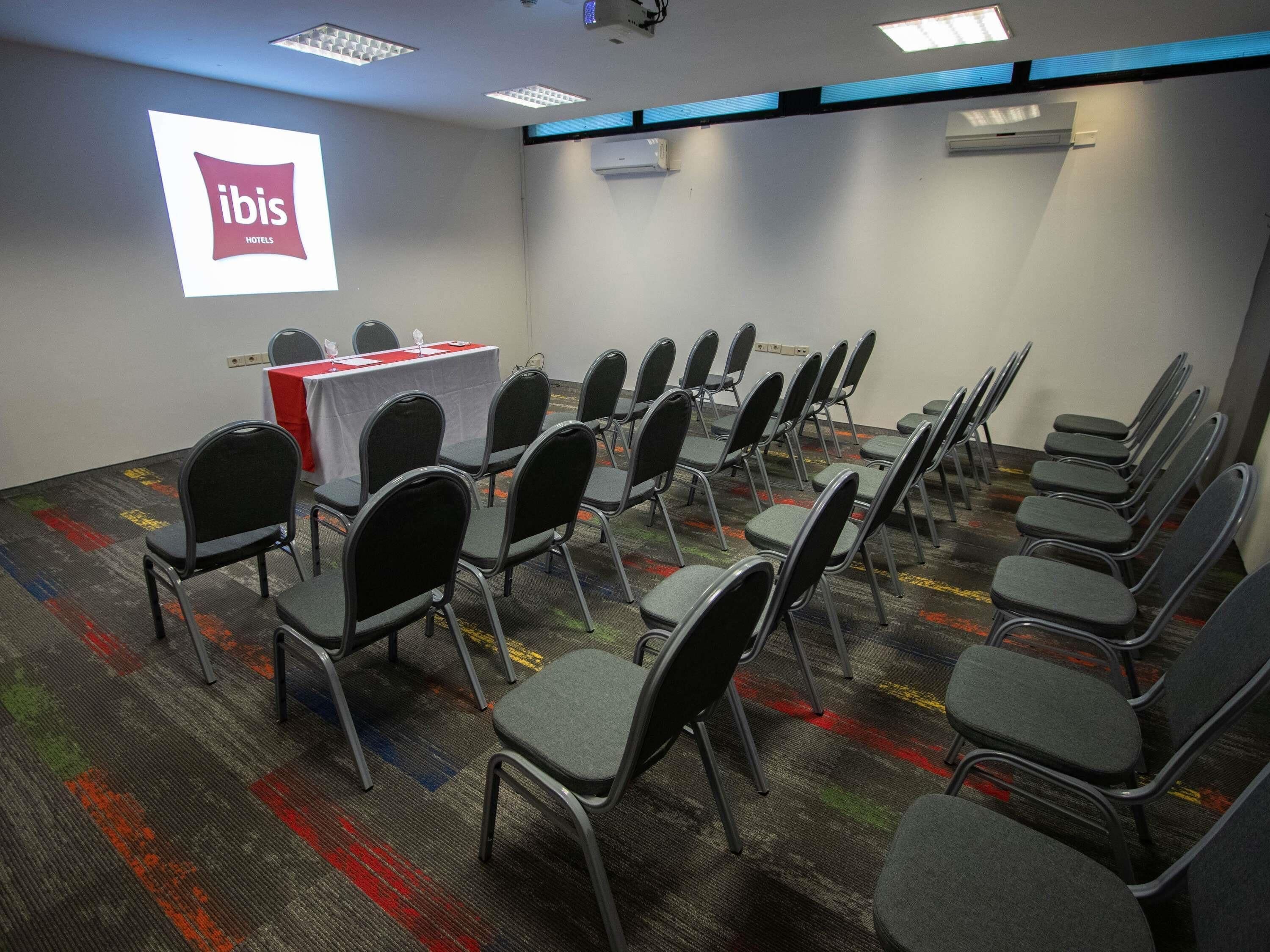 Uma sala de conferência com fileiras de cadeiras, uma mesa com toalha vermelha e uma tela de projetor exibindo o logotipo dos hotéis ibis.